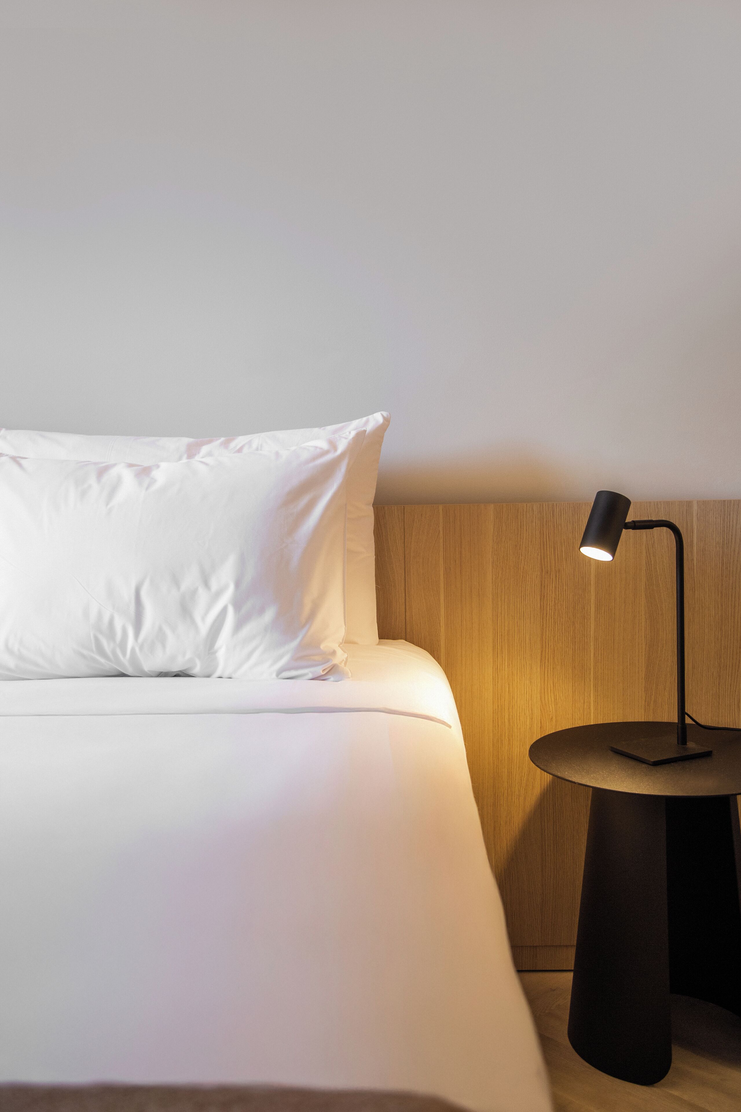 Cama bem arrumada com lençóis brancos, uma mesinha redonda com abajur preto e cabeceira de madeira.