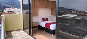 Quarto com cama, cabeceira de madeira, mesa de cabeceira e vista da cidade através de grandes janelas.
