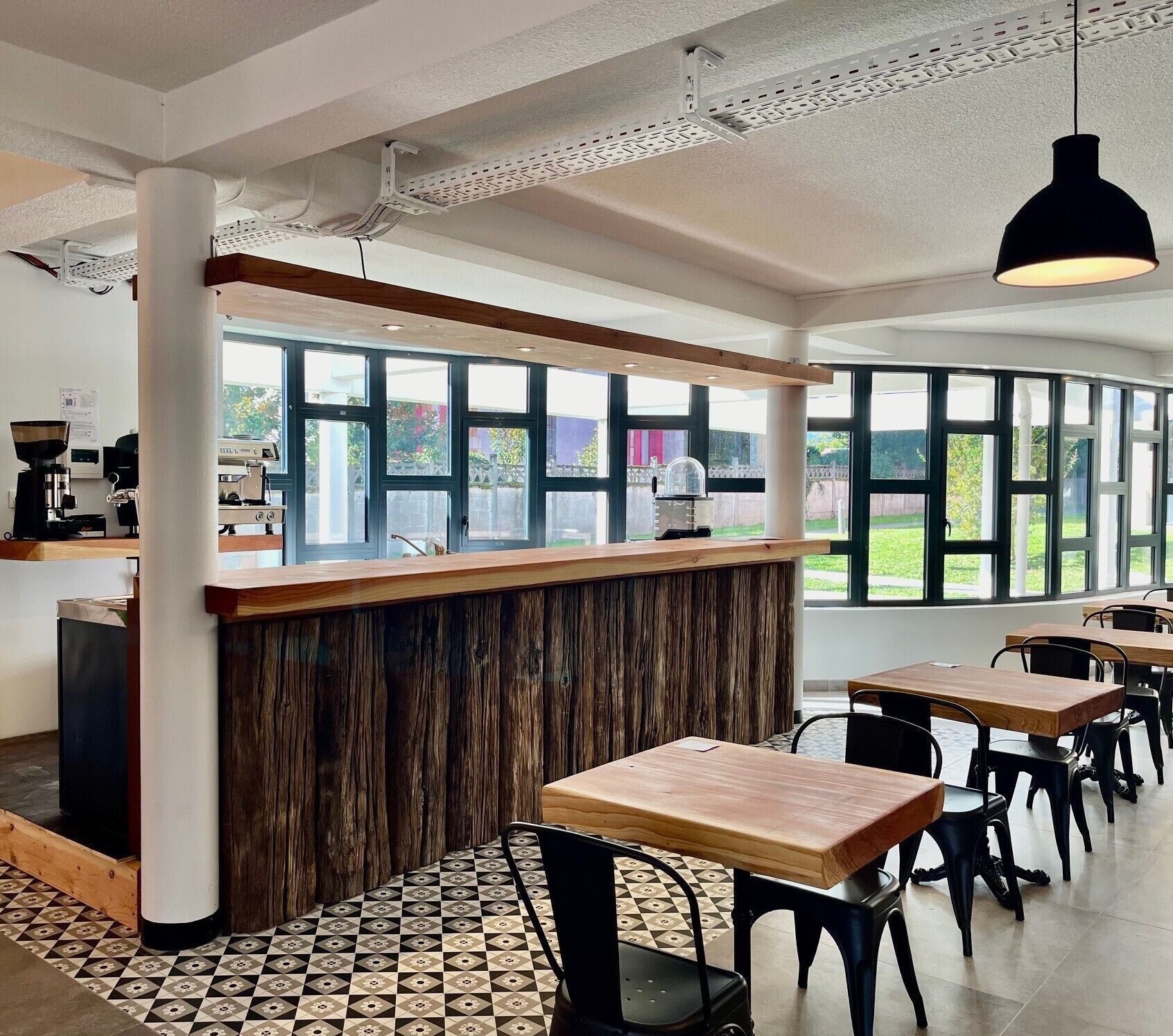 Um café moderno com balcão de madeira, cadeiras pretas e amplas janelas com vista para o exterior.