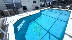 Uma piscina retangular com superfície de água azul cristalina, rodeada por paredes brancas e uma área de varanda coberta com cadeiras e mesas.