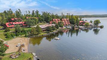 Um hotel na margem de um lago com vários edifícios de telhado vermelho, uma área de praia arenosa com espreguiçadeiras e um píer com barcos.