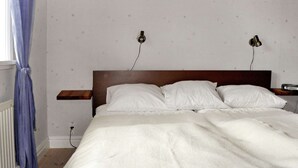 Quarto com cabeceira de madeira, duas luminárias de cabeceira, colcha branca e cortina azul.
