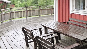 Um deck de madeira com mesa para piquenique e bancos, cercado por grades de madeira e um prédio vermelho com janela.