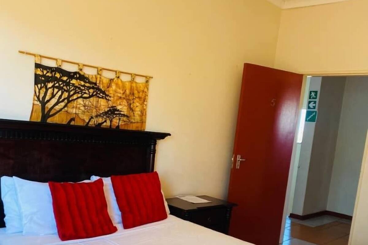 Uma cama bem arrumada com roupa de cama branca e vermelha, uma cabeceira de madeira, uma porta vermelha e uma parede com tapeçaria de árvore.