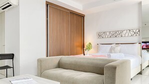 Quarto de hotel moderno com sofá, cama com roupa de cama branca, guarda-roupa de madeira, ar condicionado fixado na parede e um objeto decorativo na parede.
