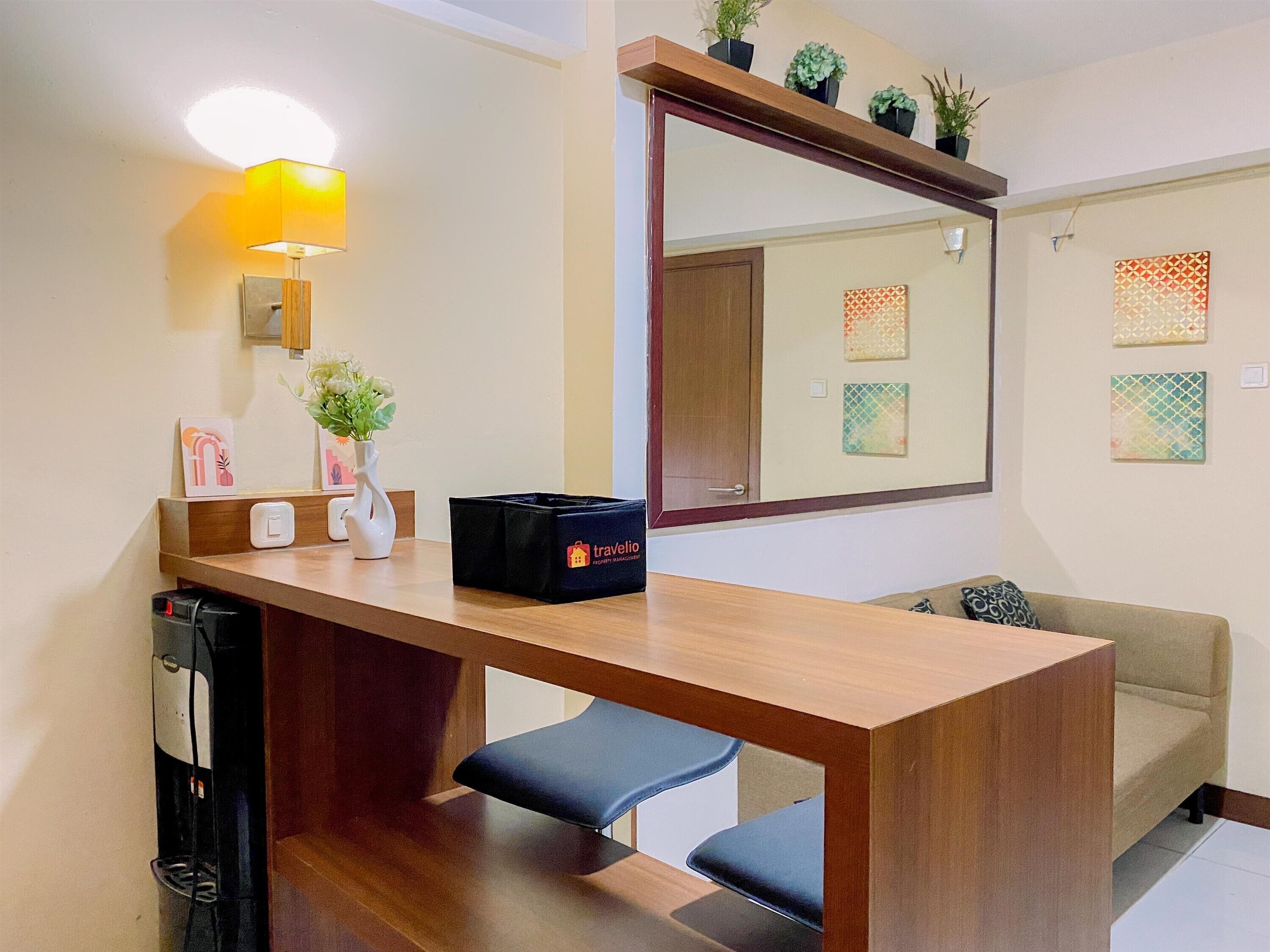 Quarto de hotel com secretária de madeira, espelho, sofá e pequeno banco. Há plantas em vasos numa prateleira acima do espelho e uma luminária na secretária.