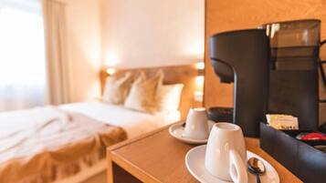 Quarto de hotel com uma cama, mesa de cabeceira com cafeteira, chávenas e uma colher.