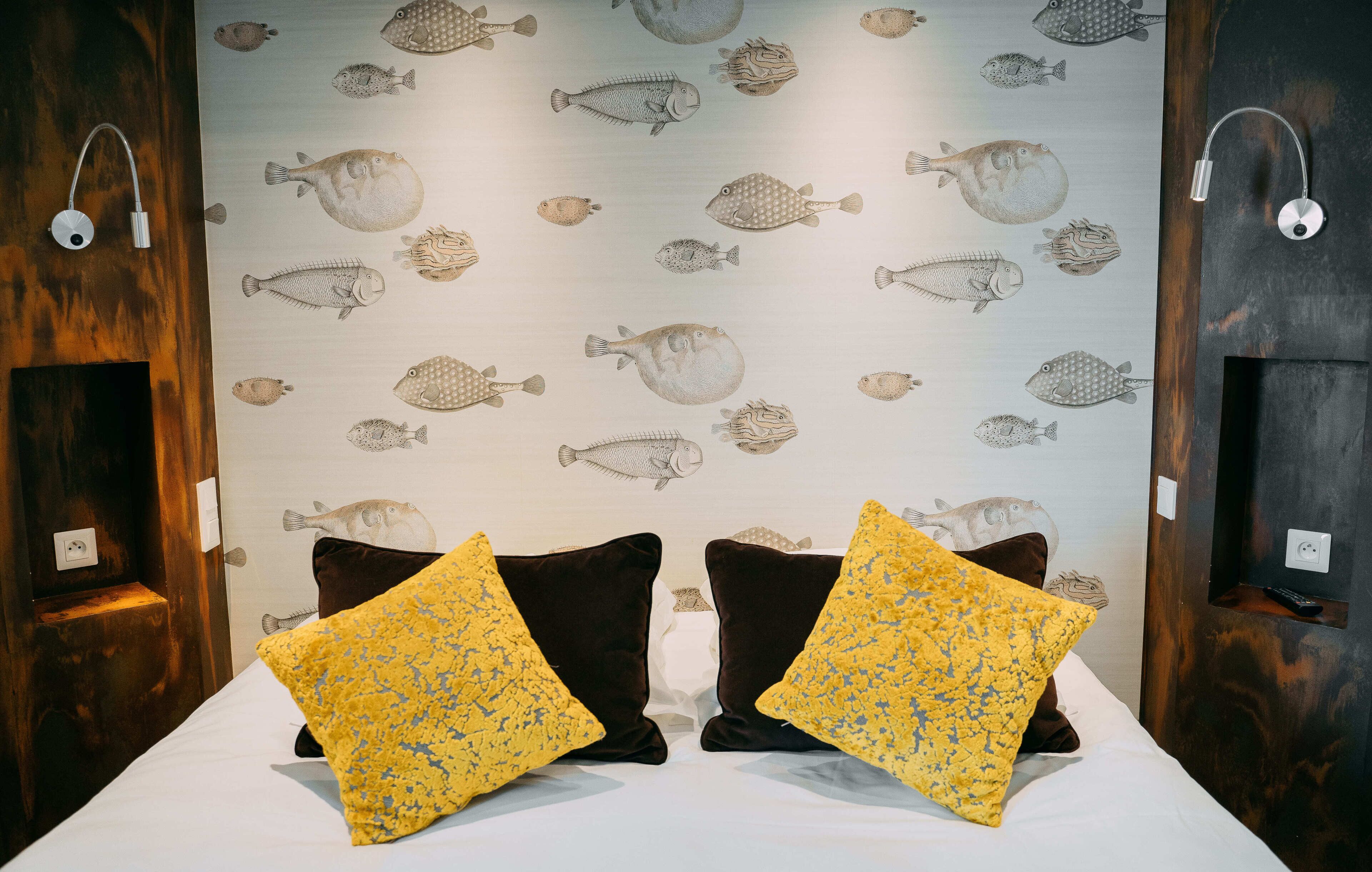 Quarto de hotel com uma cama, duas almofadas, parede com papel de parede de peixes e luminária fixada na parede.