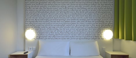 Quarto de hotel com uma cama grande, duas mesas de cabeceira e uma luminária fixada na parede.