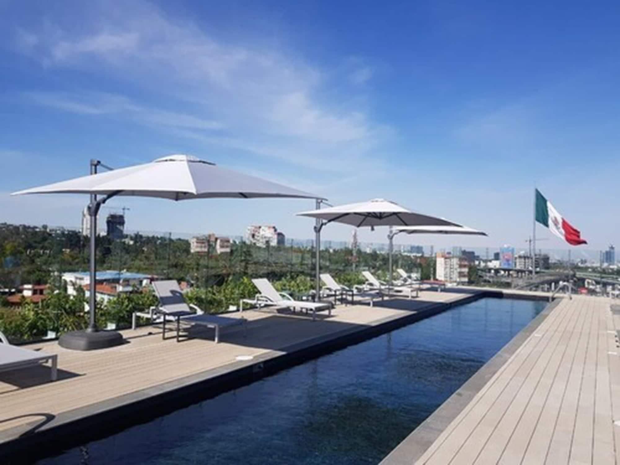 Área de piscina no terraço com espreguiçadeiras e guarda-sóis, uma bandeira e paisagem urbana ao fundo.