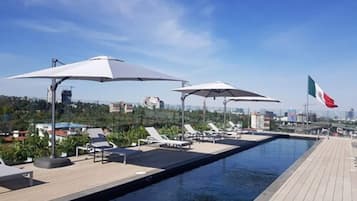 Área de piscina no terraço com espreguiçadeiras e guarda-sóis, uma bandeira e paisagem urbana ao fundo.