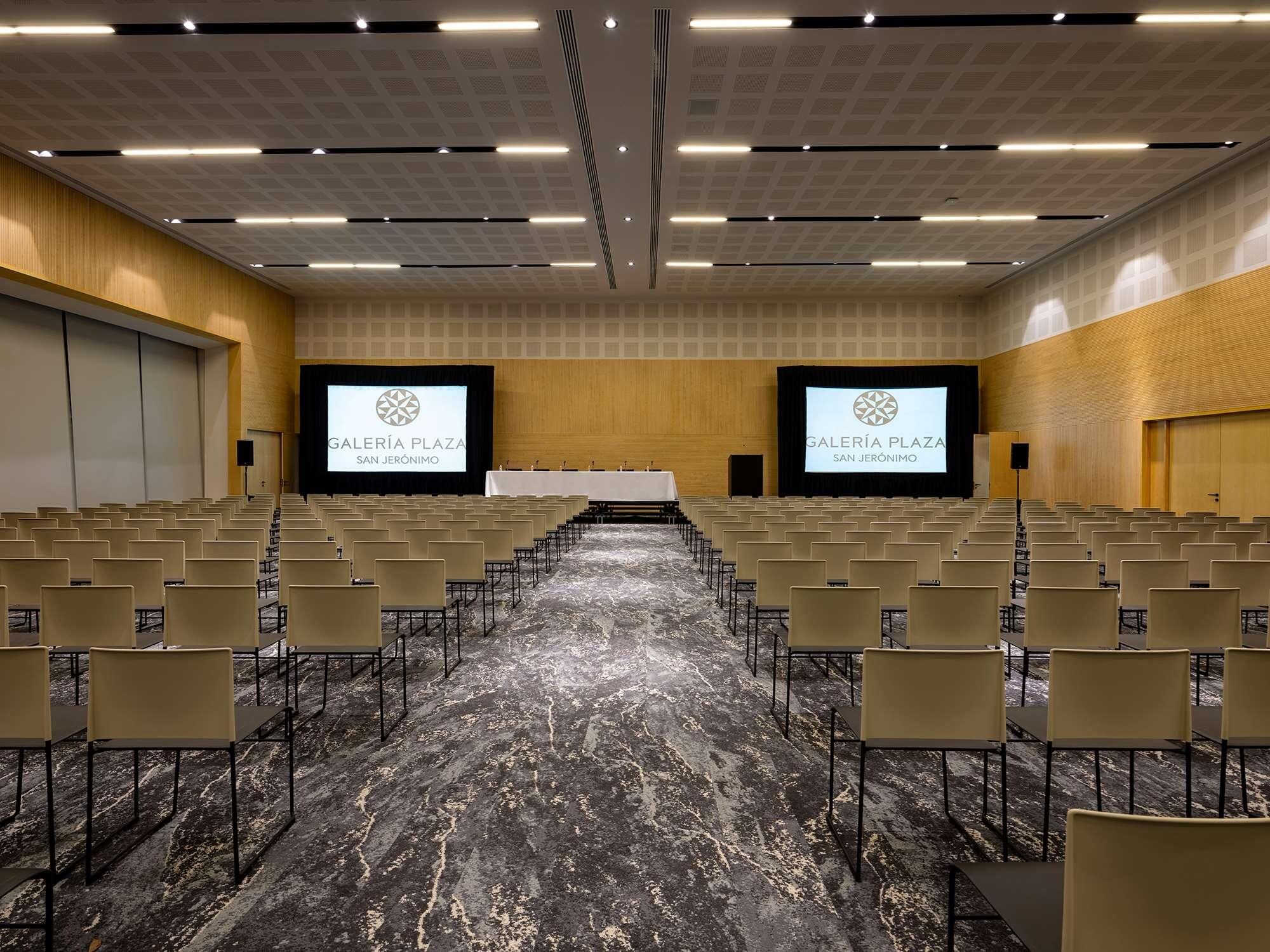 Uma sala de conferências com fileiras de cadeiras, um palco com uma tela e piso coberto de carpete.