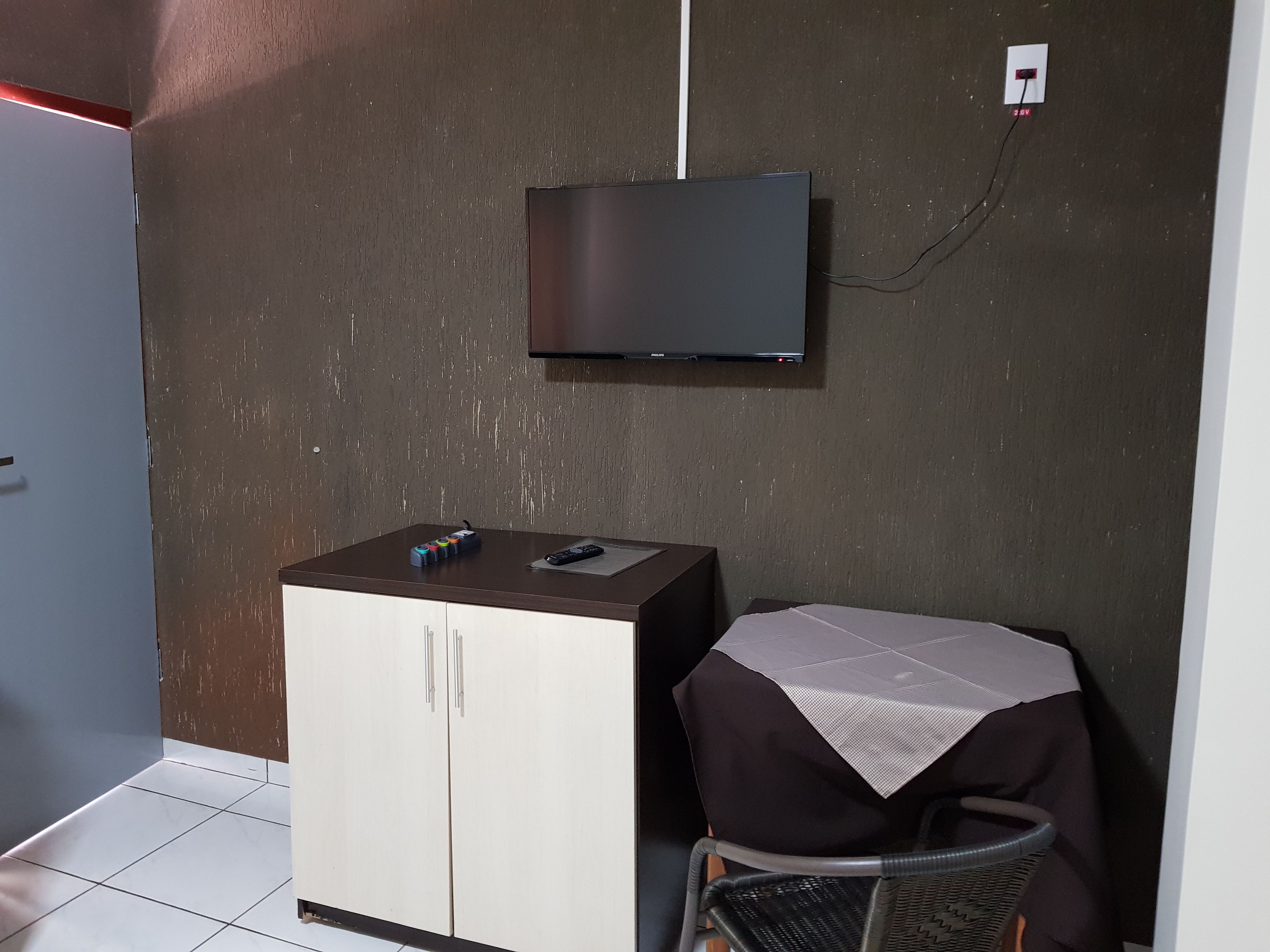 Quarto com televisão de tela plana presa na parede, um armário com gavetas, uma mesinha com toalha e uma cadeira.