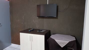 Quarto com televisão de tela plana presa na parede, um armário com gavetas, uma mesinha com toalha e uma cadeira.