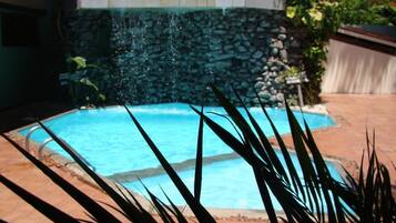 Piscina com cascata de pedra, rodeada por plantas e um terraço com ladrilhos.