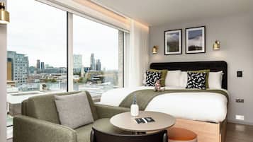 Quarto de hotel moderno com uma cama grande, um sofá, uma mesa redonda e vista para a cidade através de amplas janelas.