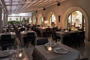 Um restaurante com várias mesas preparadas para refeições, apresentando uma área externa coberta com janelas em arco e vista para a paisagem urbana.
