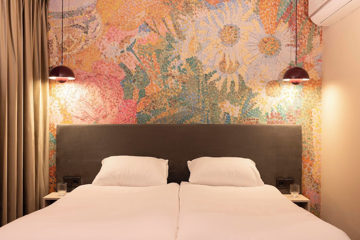 Uma cama de casal com cabeceira, duas mesas de cabeceira com luminárias e um mural floral colorido na parede.
