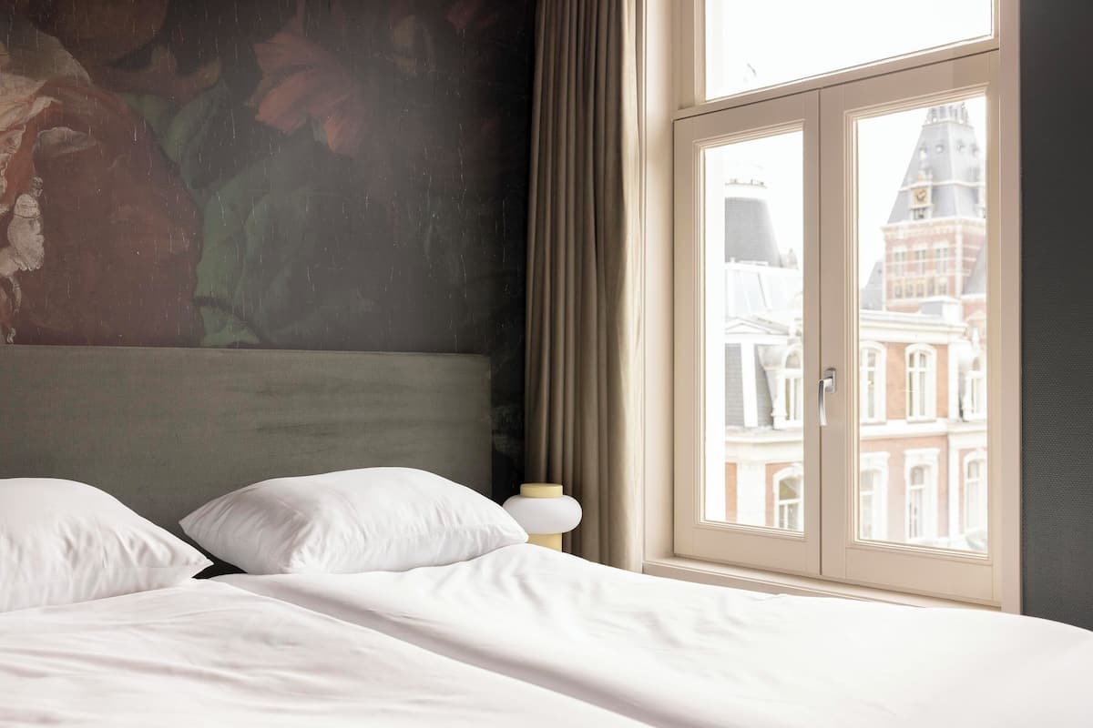 Quarto de hotel com cama, mesa de cabeceira e vista para um edifício pela janela.