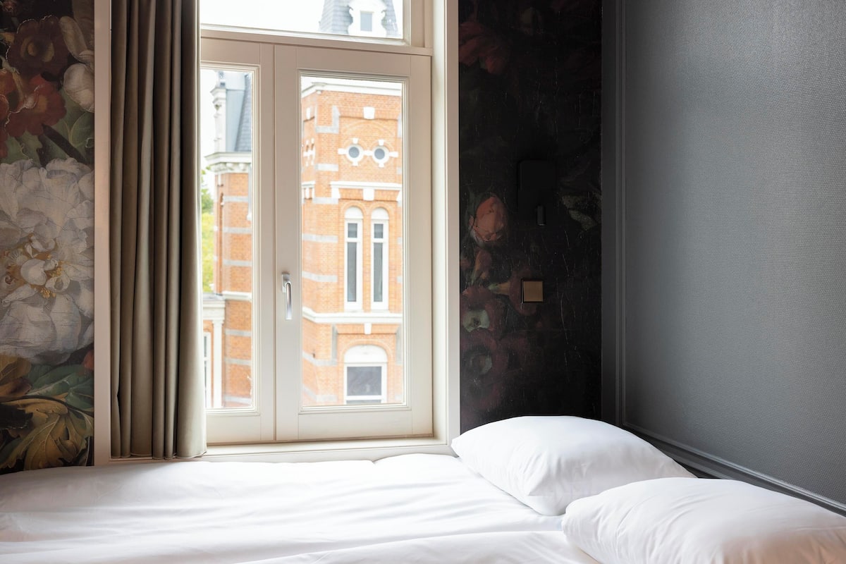 Quarto de hotel com cama, janela com cortinas e vista para um edifício de tijolos.