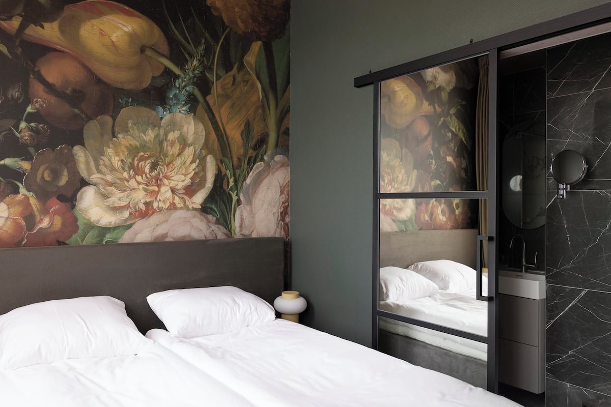 Quarto de hotel com uma cama, uma mesa de cabeceira, um espelho e um grande mural floral na parede.
