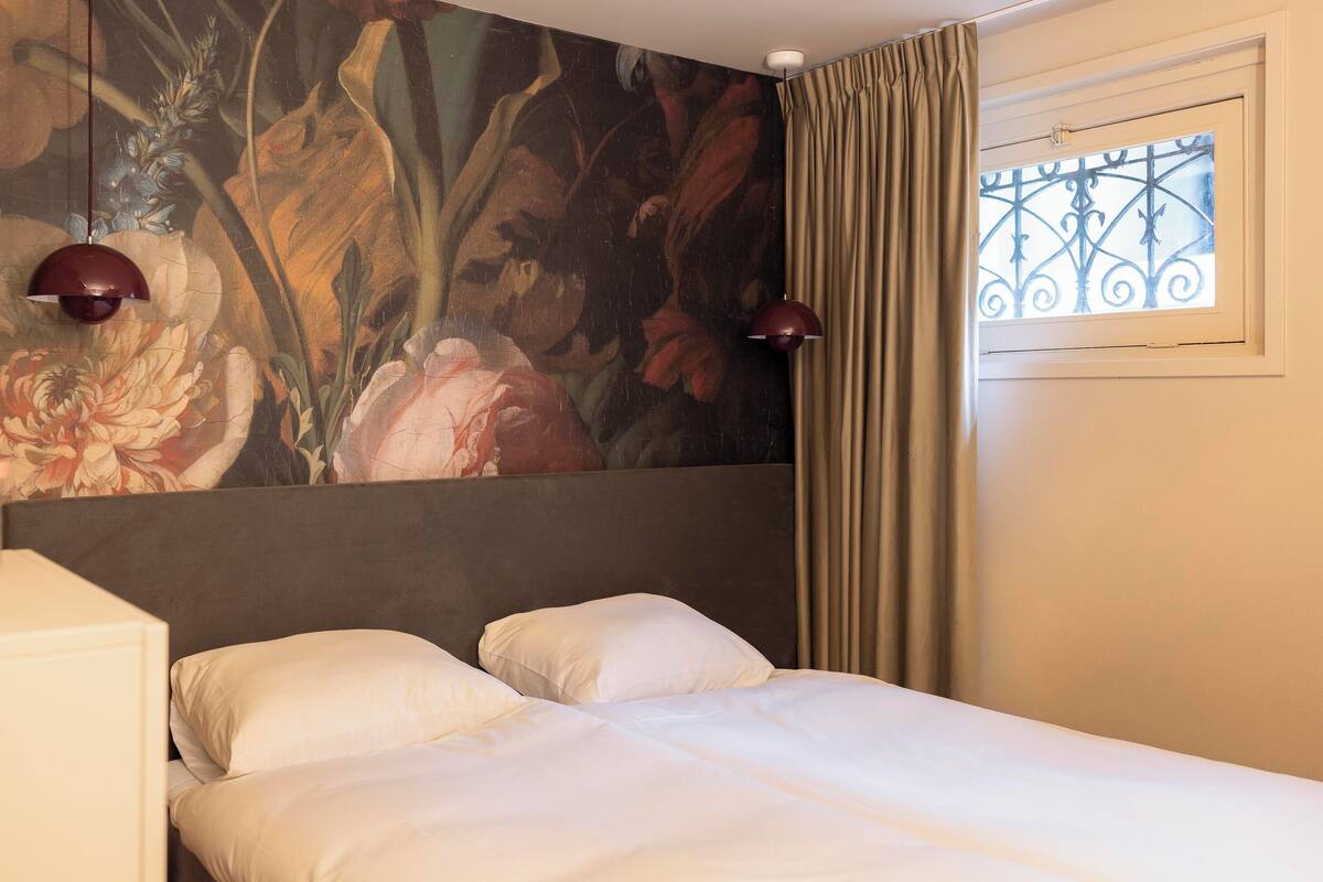Quarto de hotel com uma cama, duas luminárias, uma janela com cortinas e um mural floral na parede.
