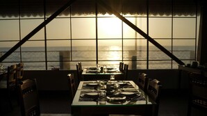 Uma sala de jantar com mesas preparadas para uma refeição, amplas janelas com vista para o mar e um teto alto com padrão em grade.