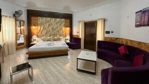 Quarto de hotel moderno com uma cama grande, área de estar com um sofá roxo e uma parede de destaque em madeira.