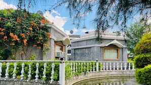 Um edifício de pedra tradicional com uma ponte curva sobre um lago, rodeado por vegetação exuberante e plantas floridas.