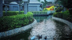 Uma corrente de água calma com uma ponte de pedra, vegetação exuberante e um edifício colorido ao fundo.