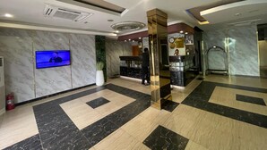 Um saguão de hotel com uma mesa de recepção, uma tela de televisão, um extintor de incêndio e uma planta.