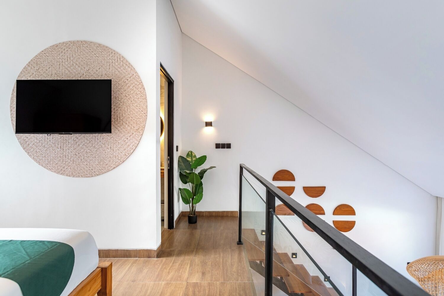 Quarto de hotel moderno com uma televisão grande fixada numa parede texturizada, uma planta em vaso e uma escada com detalhes em madeira.
