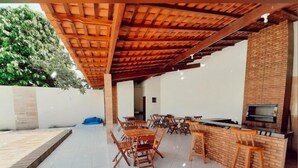 Um pátio externo com pérgola de madeira, churrasqueira de tijolos e várias mesas e cadeiras de madeira.