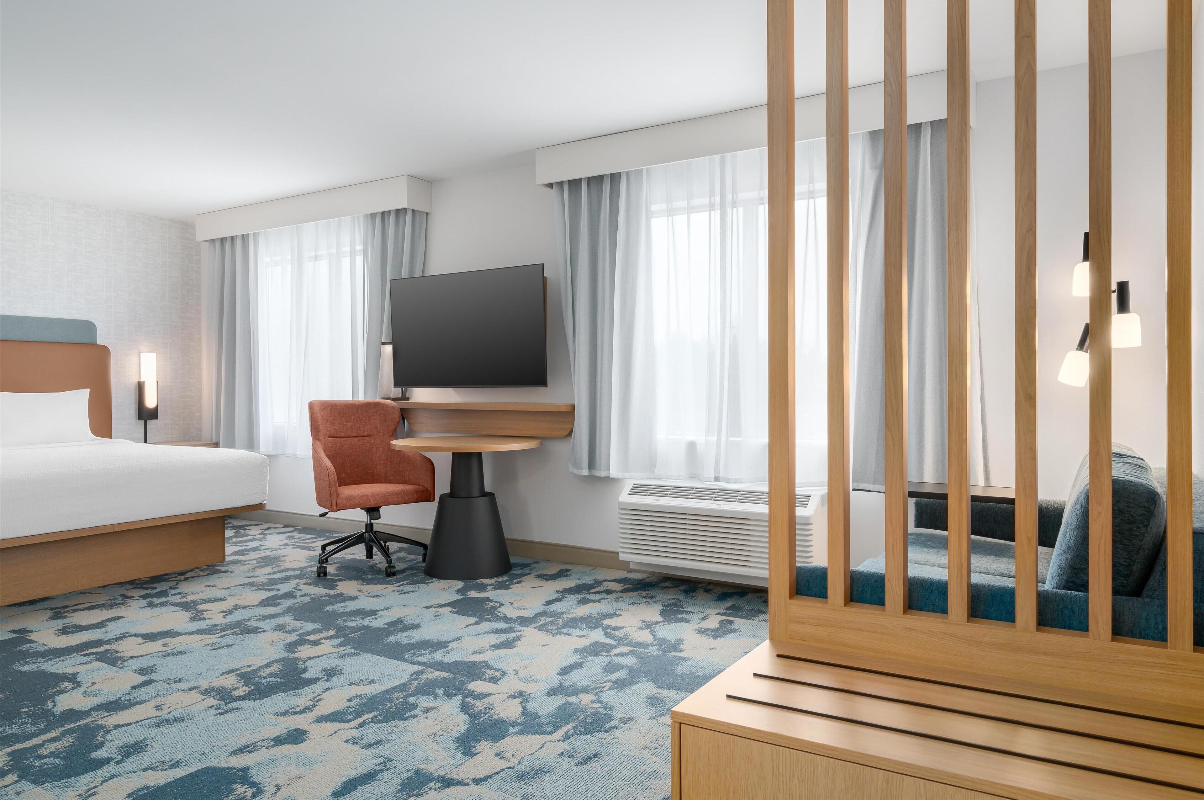 Quarto de hotel com cama, escrivaninha com cadeira, televisão de tela plana e uma grande janela com cortinas.
