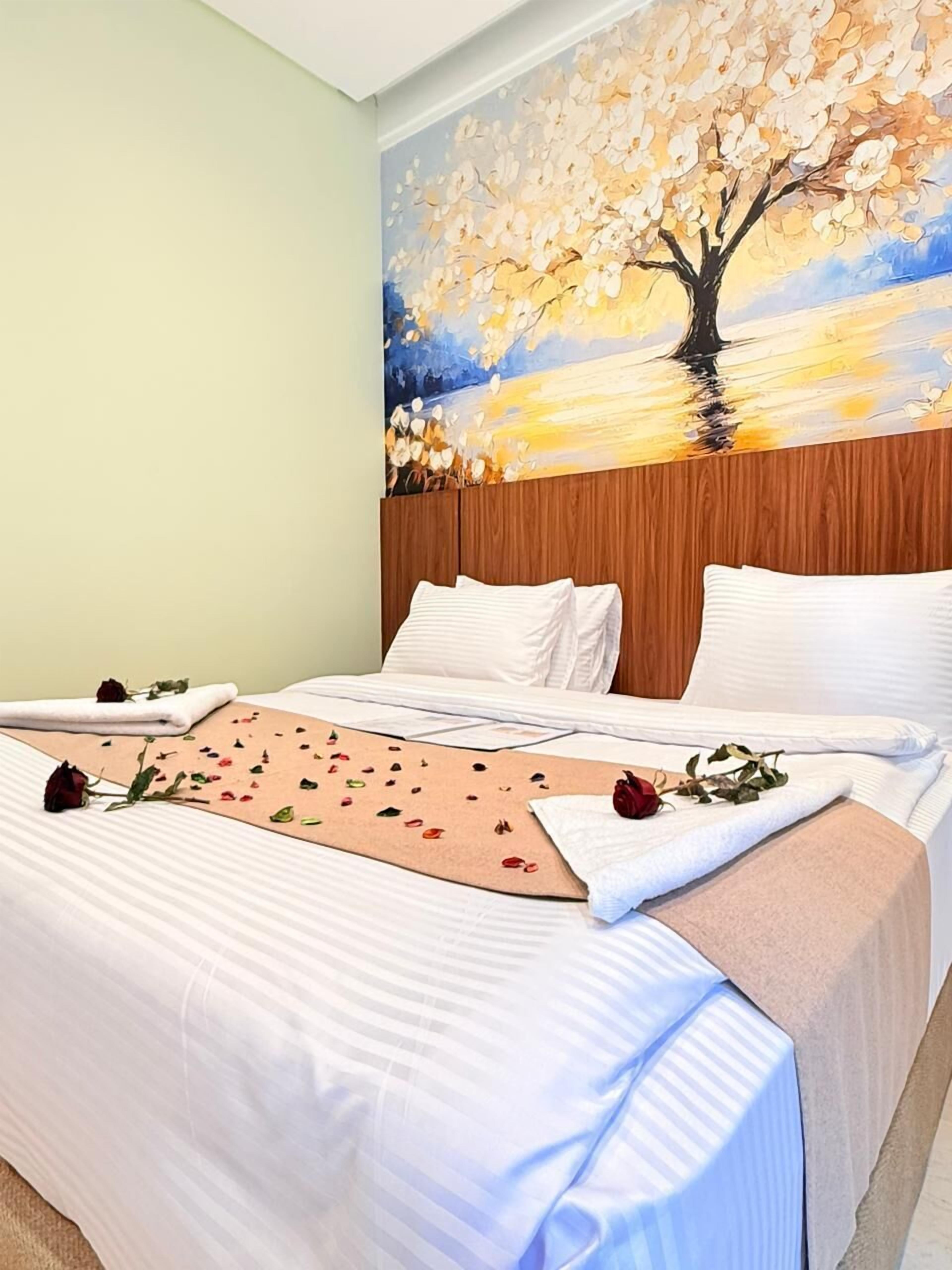 Quarto de hotel com cama, cabeceira de madeira e pintura de uma árvore com flores na parede.