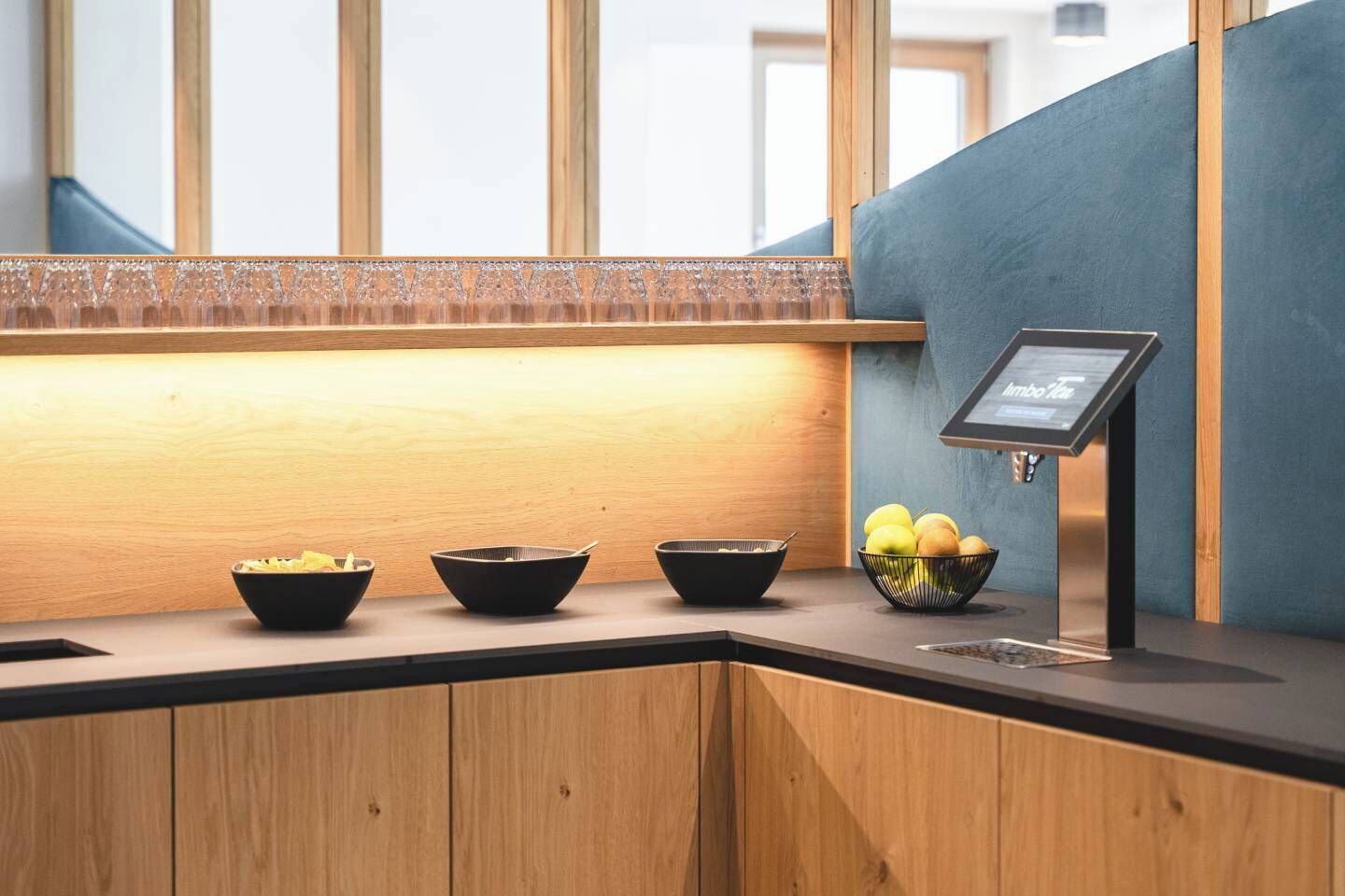 Uma área de recepção de hotel com um balcão de madeira, três recipientes com lanches, uma cesta de frutas e um painel digital de cardápio.
