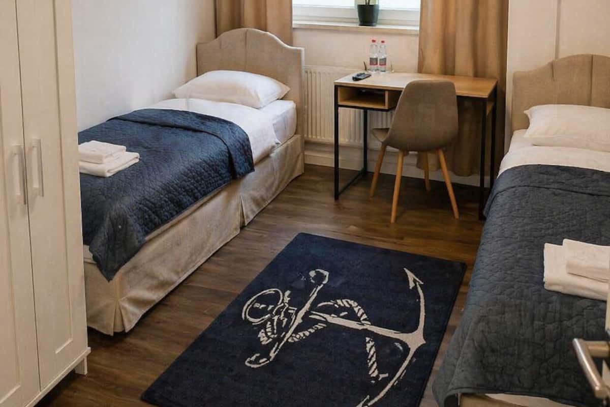 Quarto de hotel com duas camas, uma escrivaninha, uma cadeira, um guarda-roupa e uma janela com cortinas.