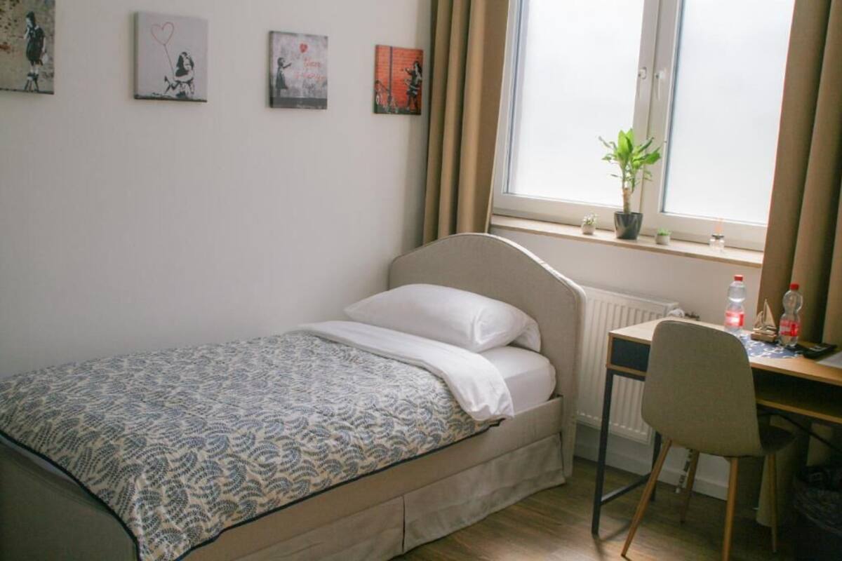 Uma cama de solteiro com um cobertor estampado, uma escrivaninha com uma cadeira, um radiador e uma janela com cortinas.