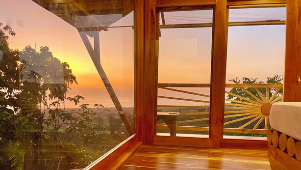 Quarto com uma grande janela com vista para o pôr do sol, piso de madeira e vista para o oceano.