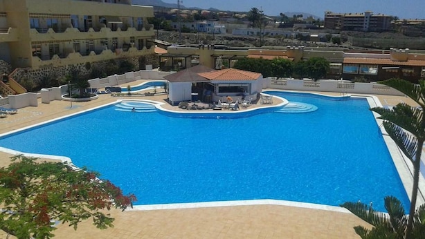 Uma piscina grande com área de estar central, rodeada por edifícios e paisagem montanhosa.