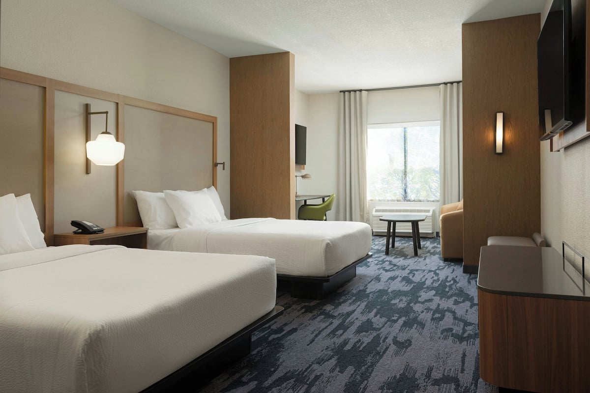 Quarto de hotel com duas camas, uma escrivaninha, uma cadeira, televisão e janela com cortinas.