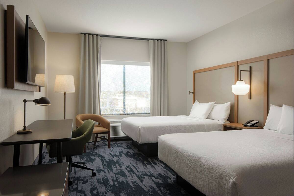 Quarto de hotel com duas camas, uma escrivaninha, uma cadeira, uma televisão e uma janela com cortinas.