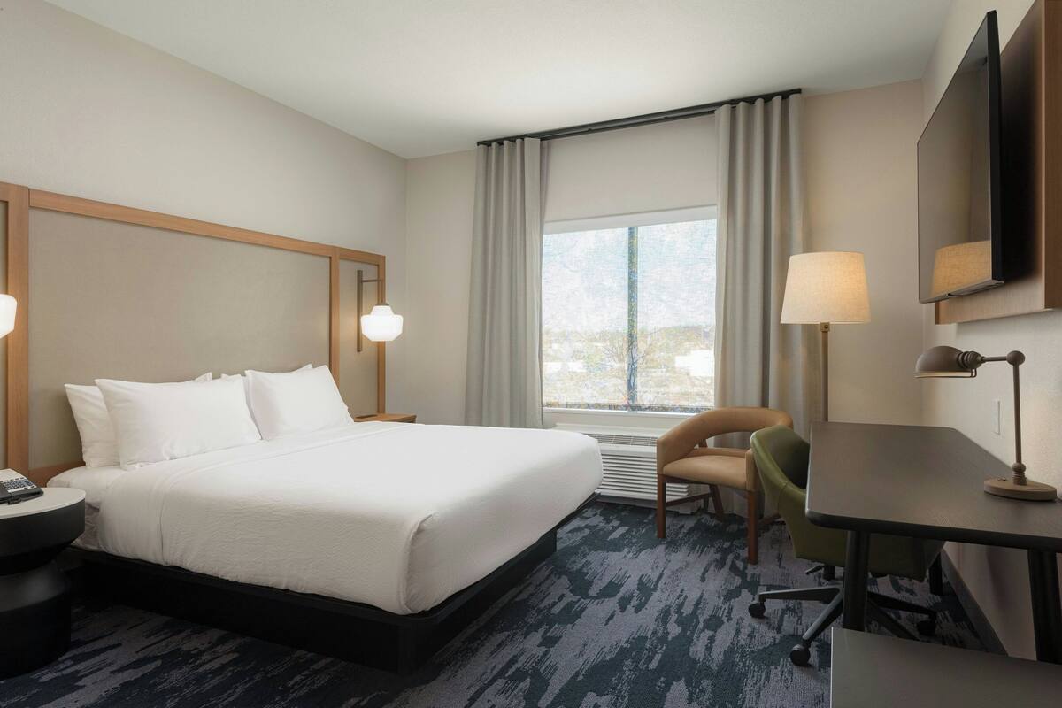 Quarto de hotel com cama, escrivaninha, cadeira, televisão e janela com cortinas.