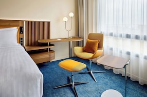 Quarto de hotel com cama, escrivaninha com luminária, poltrona amarela e uma pequena mesa.