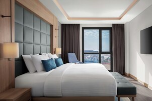 Quarto de hotel moderno com uma cama grande, área de estar e vista para a cidade pela janela.
