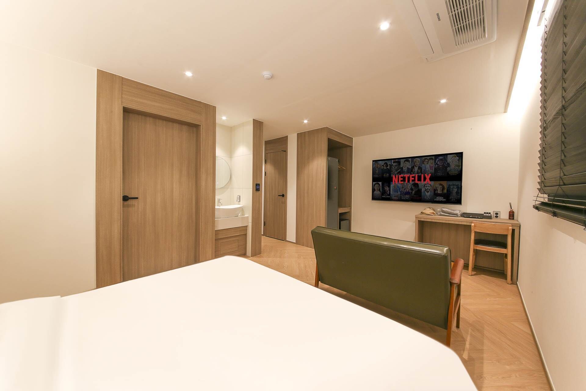 Quarto de hotel com cama, escrivaninha com cadeira, televisão na parede e banheiro visível por uma porta aberta.
