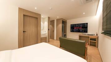 Quarto de hotel com cama, escrivaninha com cadeira, televisão na parede e banheiro visível por uma porta aberta.
