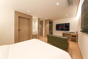 Quarto de hotel com cama, escrivaninha com cadeira, televisão na parede e banheiro visível por uma porta aberta.