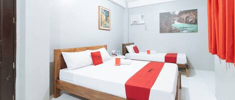 Quarto de hotel com duas camas, roupa de cama branca, detalhes em vermelho, um quadro na parede e uma janela com unidade de ar condicionado.