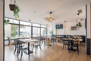 Um restaurante espaçoso com mesas e cadeiras de madeira, janelas amplas, plantas pendentes e luminárias modernas.